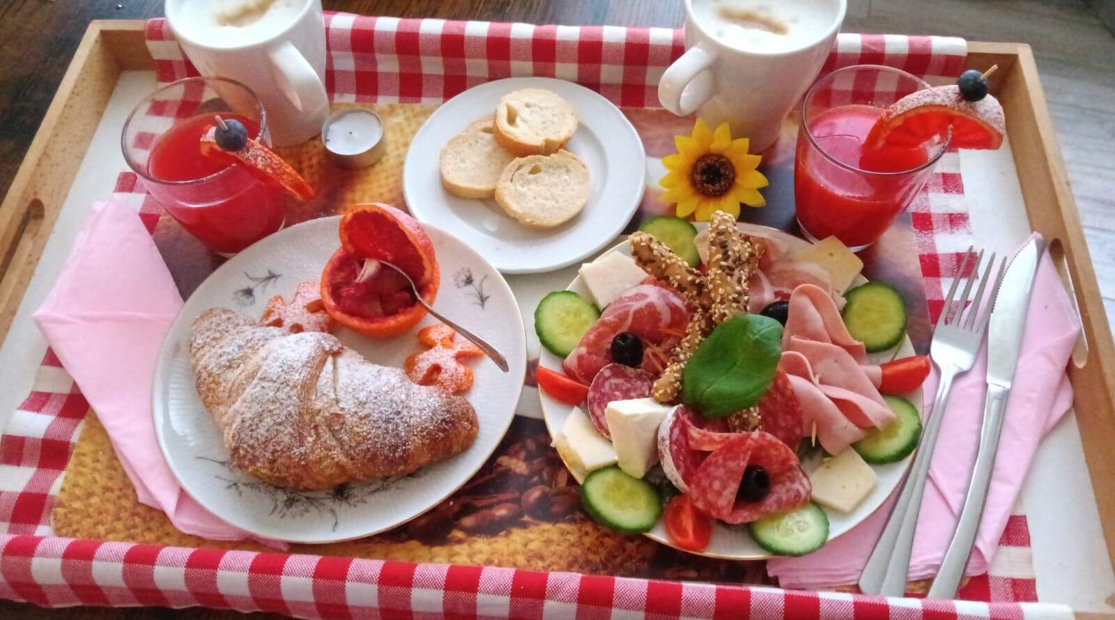Uma bandeja de café da manhã com croissant, pão fatiado, queijo, presunto, salame, azeitonas, pepino e um girassol. Há também dois copos de bebida vermelha e uma pequena vela.