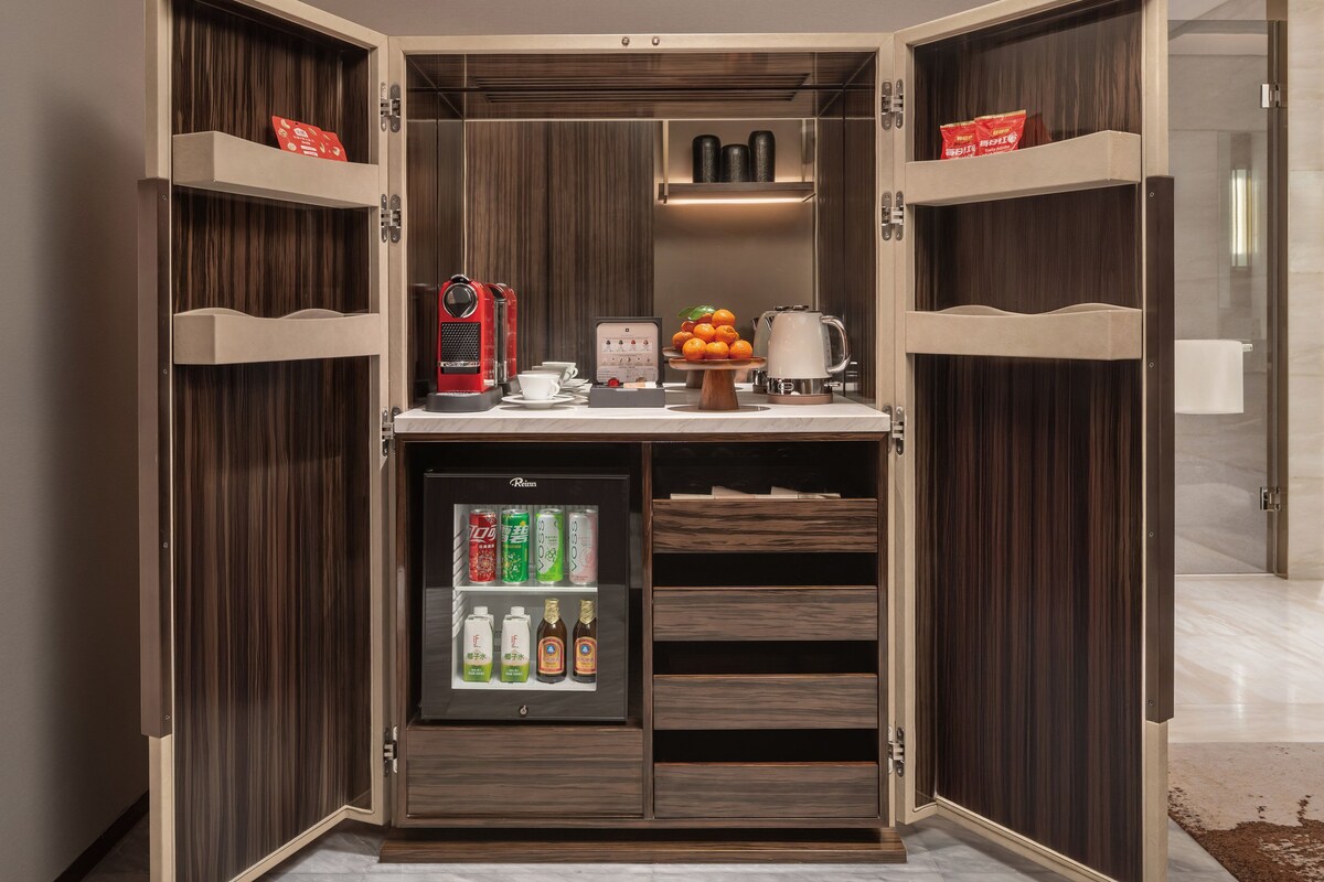 Minibar compacto com frigorífico, máquina de café e pequena seleção de bebidas.