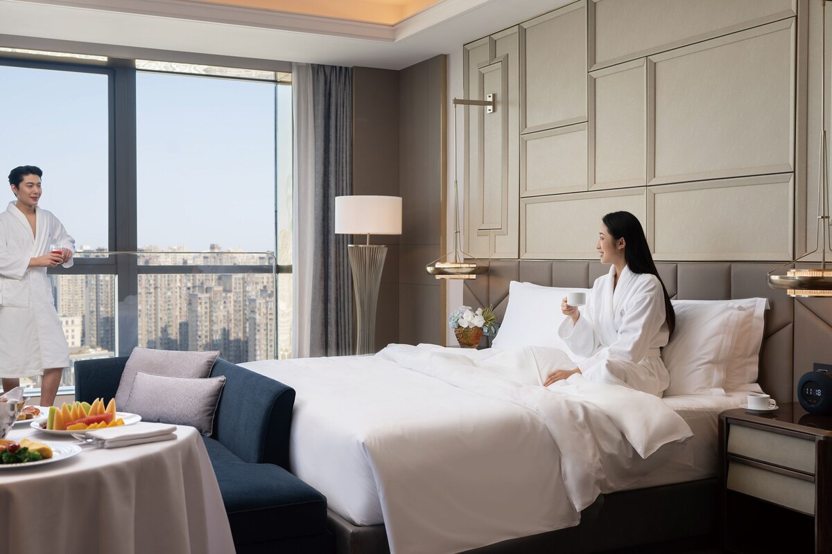 Quarto de hotel com uma cama grande, mesa de cabeceira com luminária, criado-mudo com relógio e vista da cidade através de amplas janelas.
