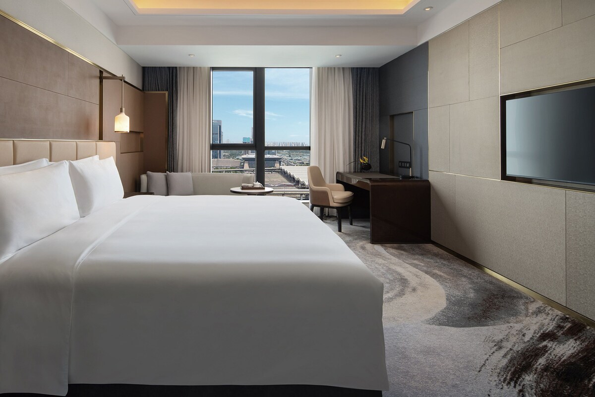 Quarto de hotel com uma cama grande, uma escrivaninha, uma cadeira, televisão e vista para a paisagem urbana.