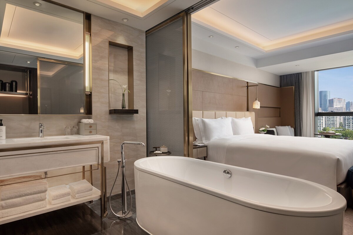 Uma casa de banho de hotel moderna com uma banheira grande, uma toucador com espelho e uma cama com vista para a cidade.
