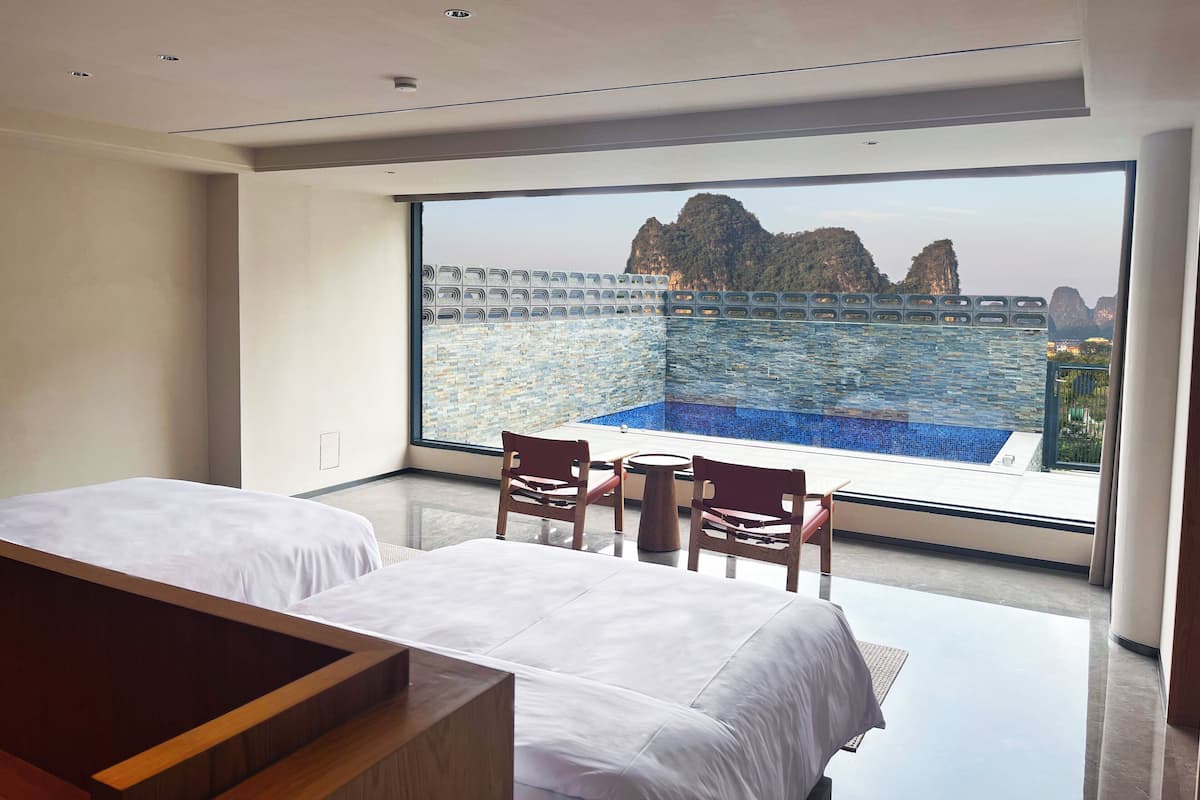 Quarto de hotel com duas camas, uma mesinha e cadeiras. Possui uma janela grande com vista para uma piscina e montanhas.