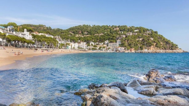 Uma praia com água cristalina, costa rochosa e edifícios numa encosta.