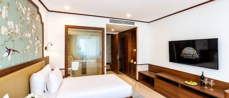 Quarto de hotel com cama, televisão, escrivaninha e banheiro visível por uma porta de vidro.