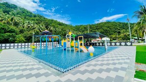 Piscina com escorrega e estruturas de recreação, cercada por um deck ladrilhado e palmeiras.