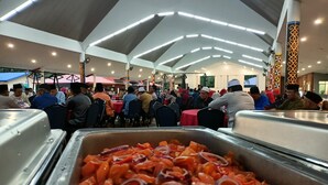 Uma área de buffet com diversos tipos de comida, incluindo legumes e carnes fatiadas, sob uma cobertura com várias mesas e cadeiras.