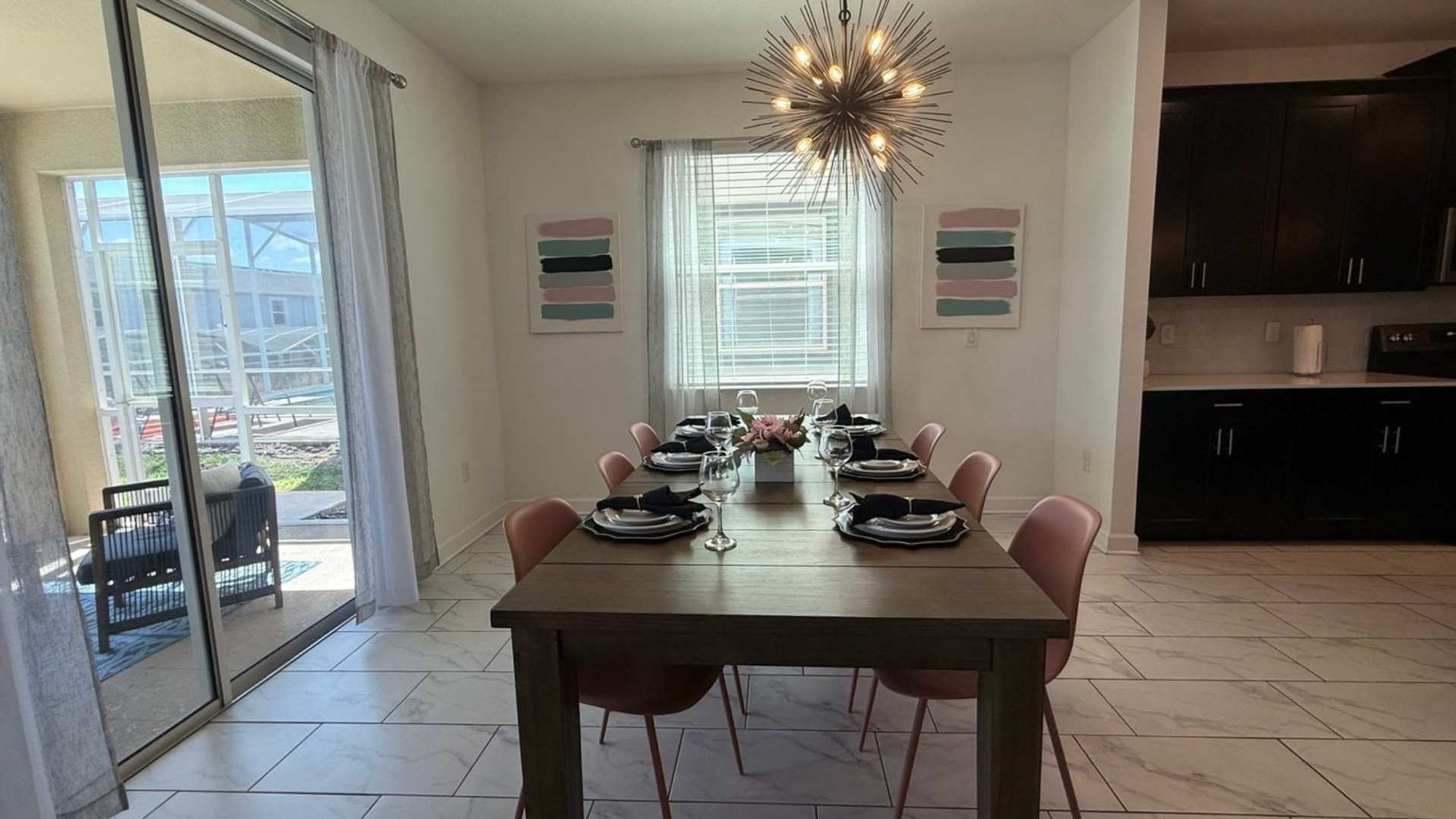 Uma área de jantar com uma mesa posta para seis pessoas, um lustre moderno e vista para uma área externa através de uma porta de vidro.