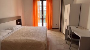Quarto de hotel com uma cama, uma escrivaninha e uma cadeira. Há uma janela com cortinas laranja e uma porta que dá para outro quarto.