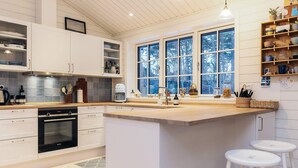 Cozinha moderna com armários brancos, uma ilha grande com banquetas, cafeteira e vista para árvores através de amplas janelas.