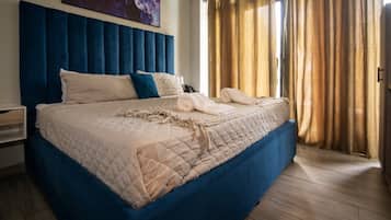 Uma cama grande com cabeceira azul, colcha acolchoada bege e manta branca.