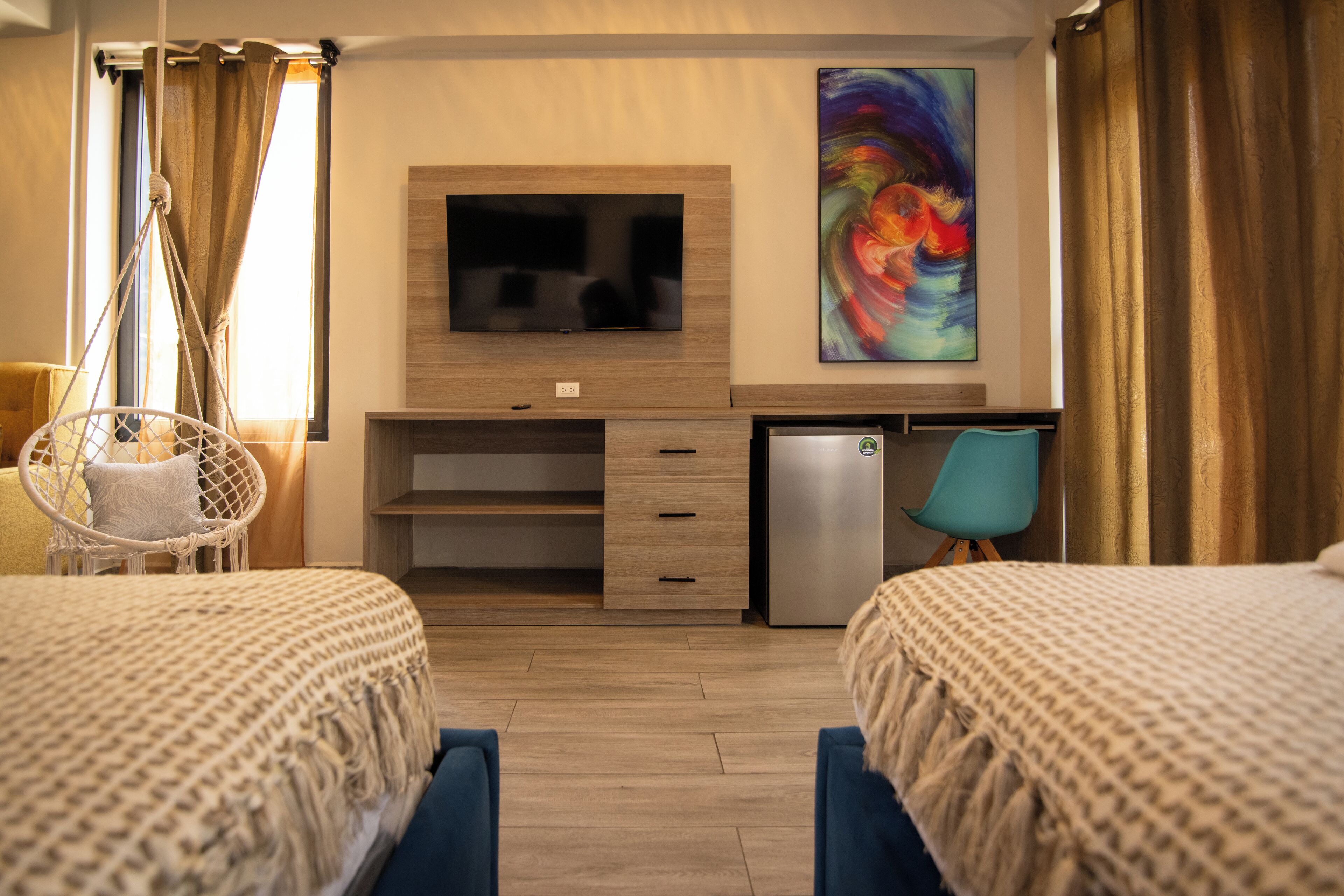 Quarto de hotel com duas camas, televisão, escrivaninha com computador, frigorífico e um quadro colorido na parede.
