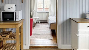 Um quarto acolhedor com piso de madeira, uma cama com colcha estampada, uma cadeira branca e um micro-ondas sobre uma mesa de madeira.