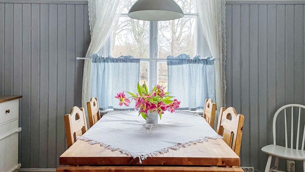 Uma sala de jantar com uma mesa de madeira, cadeiras, um vaso com flores e uma janela com cortinas.