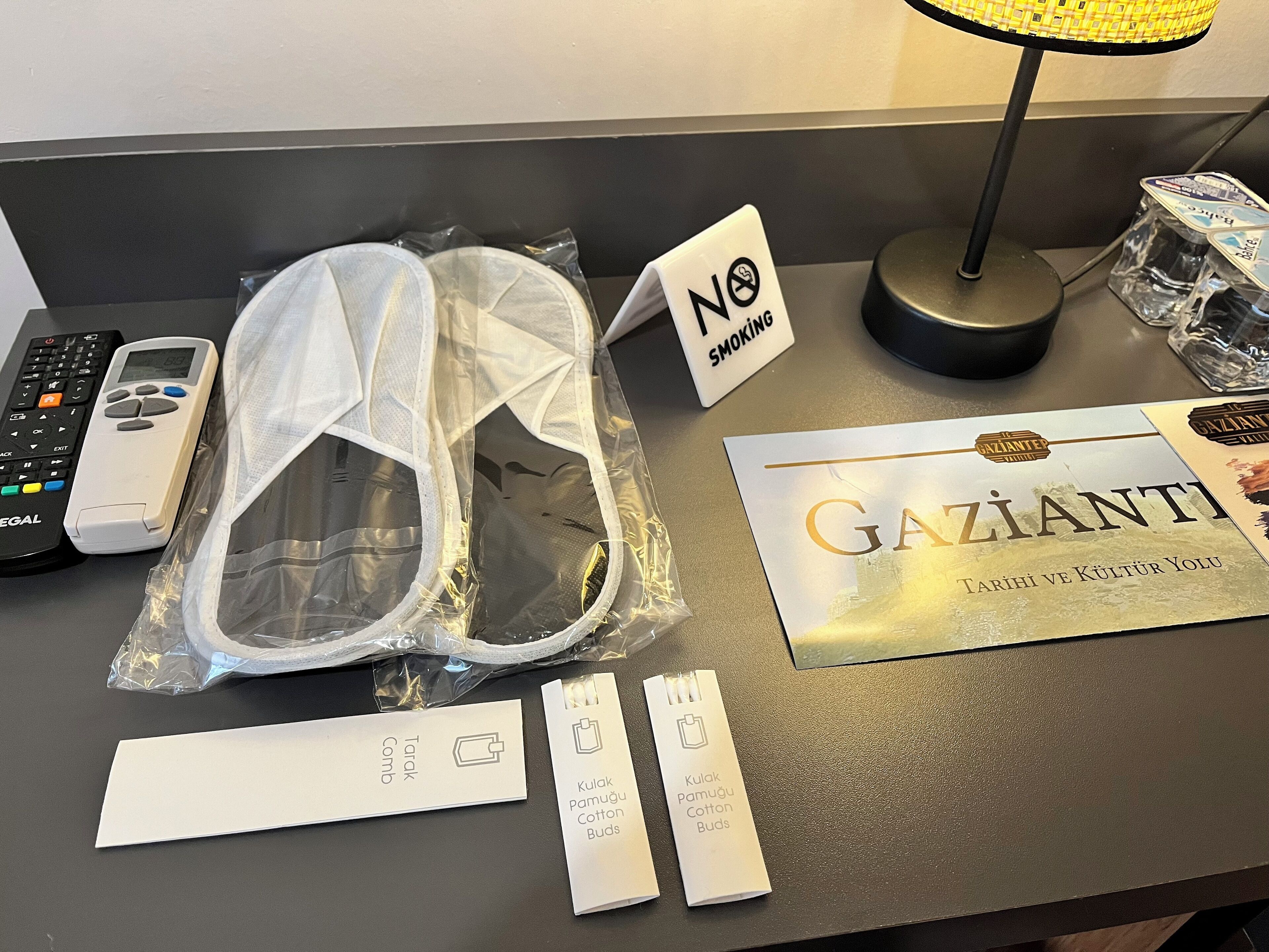 Bancada de recepção de hotel com aviso de proibido fumar, um controle remoto, fones de ouvido, um folheto e alguns itens embalados.