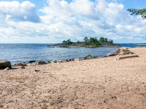 Uma praia arenosa com pedras espalhadas e uma pequena ilha com árvores ao fundo.