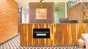 Um balcão de recepção com uma placa indicando "TOWNHOUSE" e a inscrição "Serviço da Empresa". Há um relógio na parede e um ventilador de teto acima.