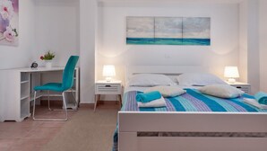 Quarto de hotel com uma cama, duas mesas de cabeceira, uma escrivaninha com cadeira, um quadro na parede e uma pequena planta.