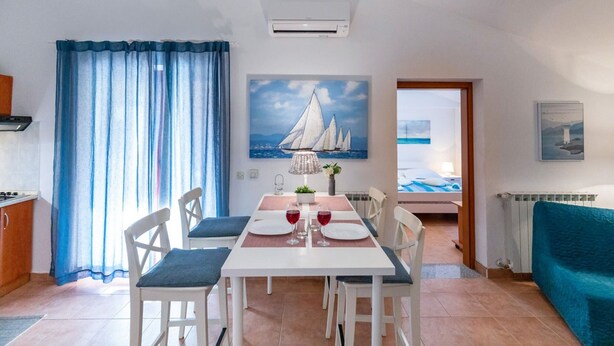 Uma área de refeições com uma mesa posta para dois, uma pintura de barcos à vela e vista para o mar através de uma janela.