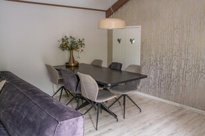 Uma área de refeições com mesa escura, cadeiras cinzentas, um vaso com flores e um lustre.