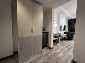 Quarto de hotel moderno com piso de madeira, guarda-roupa com linhas verticais, pequena mesa de trabalho com cadeira, sofá e televisão na parede.