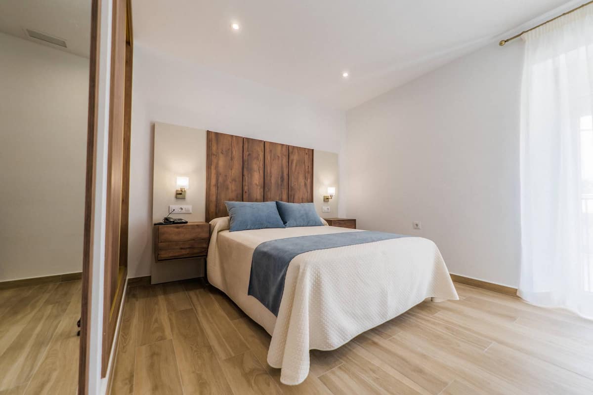 Quarto com cama, mesas de cabeceira, cabeceira de madeira e janela com cortinas.