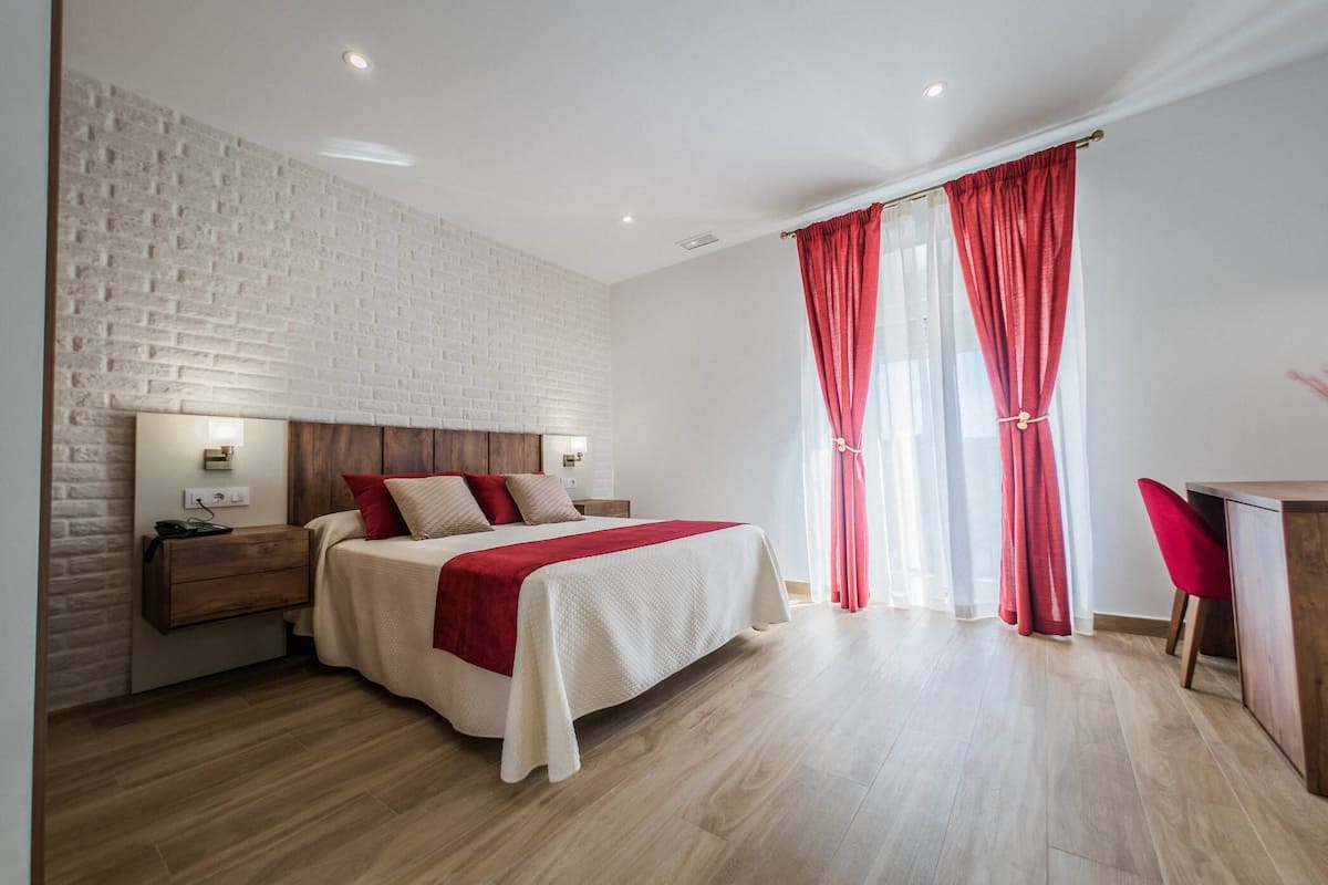 Quarto moderno com parede de tijolos, cabeceira de madeira, cama com roupa de cama vermelha e branca, mesa de cabeceira, escrivaninha com cadeira e janela com cortinas vermelhas.