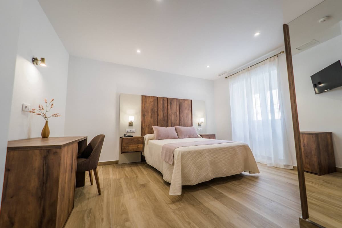 Quarto moderno com cabeceira de madeira, cama com colcha bege, escrivaninha com cadeira e televisão de tela plana na parede.