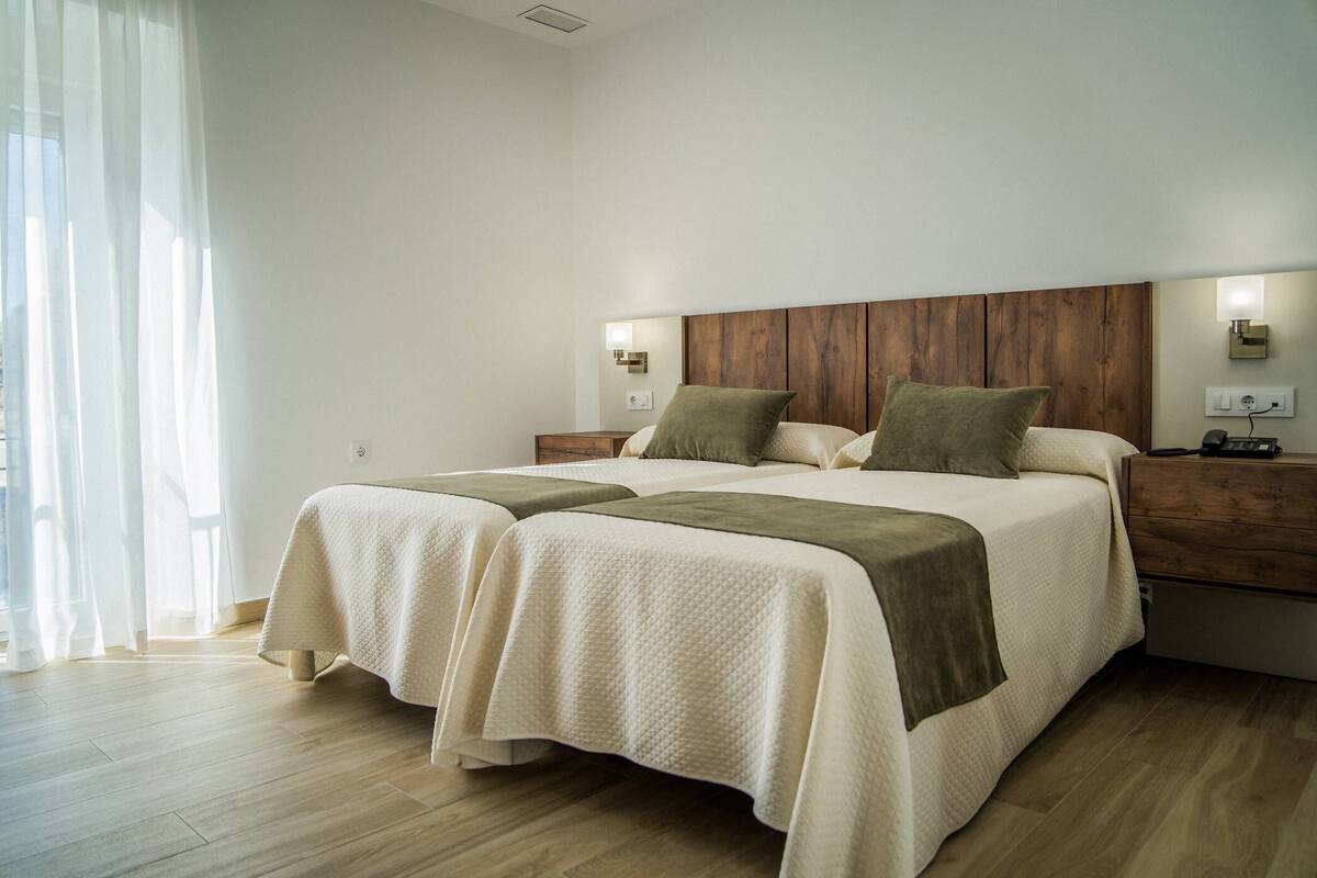 Uma cama de casal com cabeceira de madeira, duas mesas de cabeceira e uma janela grande com cortinas leves.