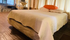 Uma cama bem arrumada com edredom branco e travesseiro laranja, uma escrivaninha pequena com um computador e uma janela com cortinas.