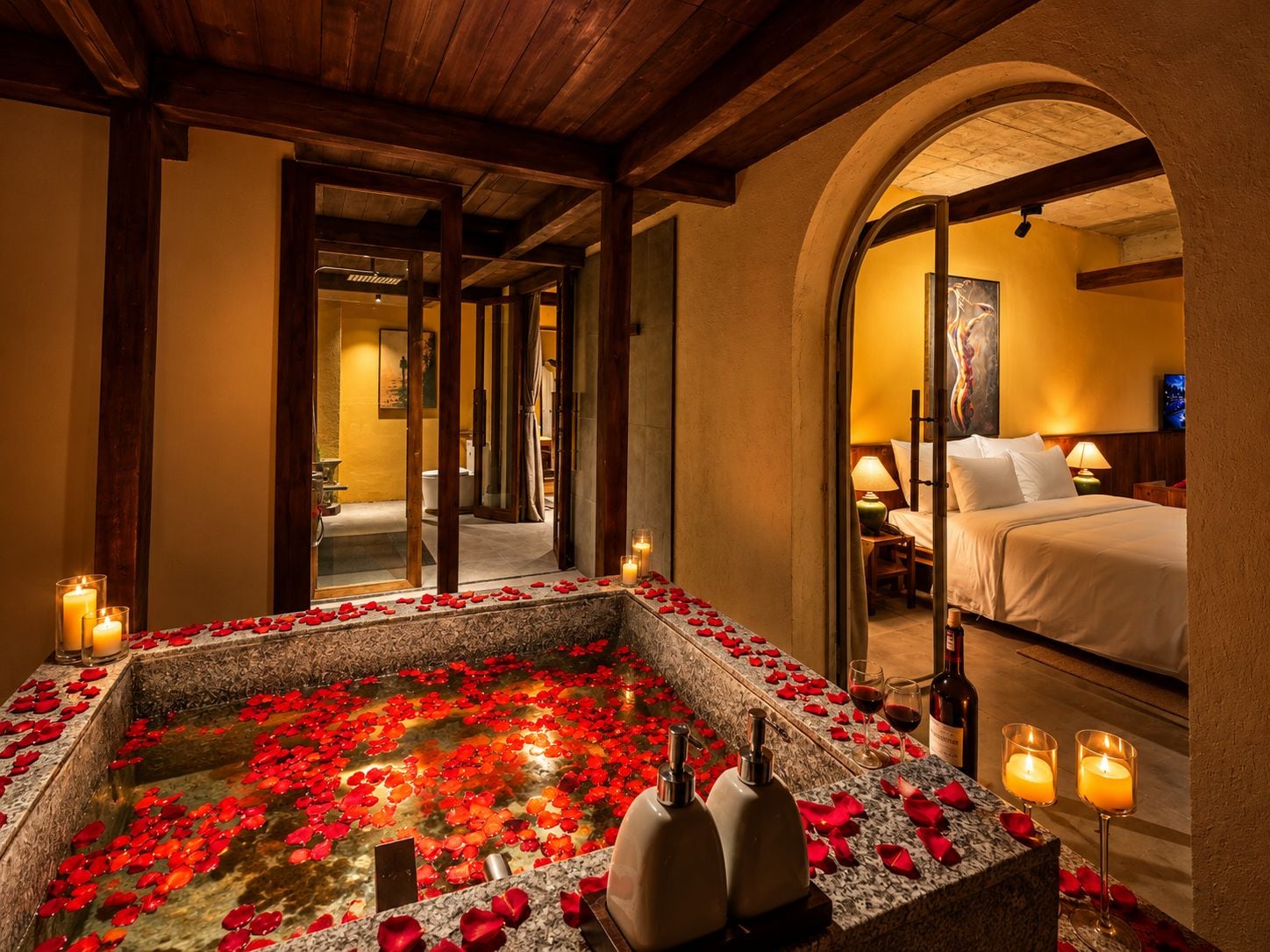 Uma área de spa com hidromassagem, rodeada por pétalas de rosa, velas e uma cama ao fundo.