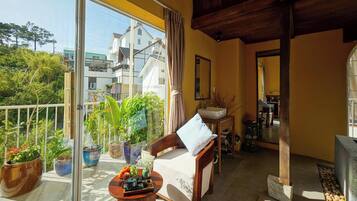 Varanda com plantas em vasos, uma cadeira de madeira, uma mesa pequena com uma chaleira e vista para edifícios e árvores.