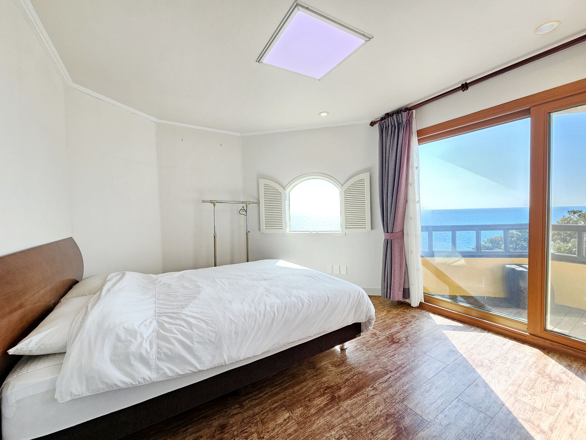 Quarto com cama, claraboia, janela com persianas e varanda com vista para o mar.