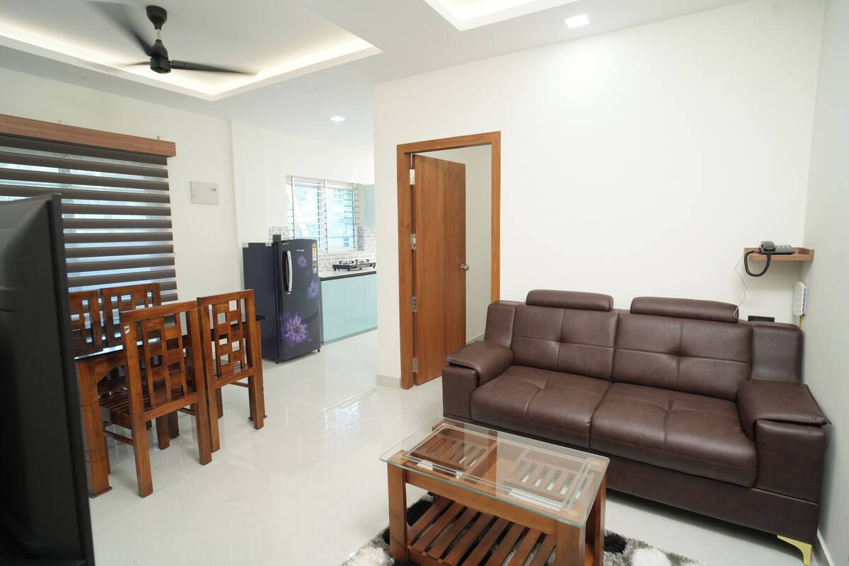 Uma sala de estar com sofá de couro marrom, mesa de jantar de madeira com cadeiras, mesa de centro de vidro e um ventilador de teto, com uma cozinha ao fundo.