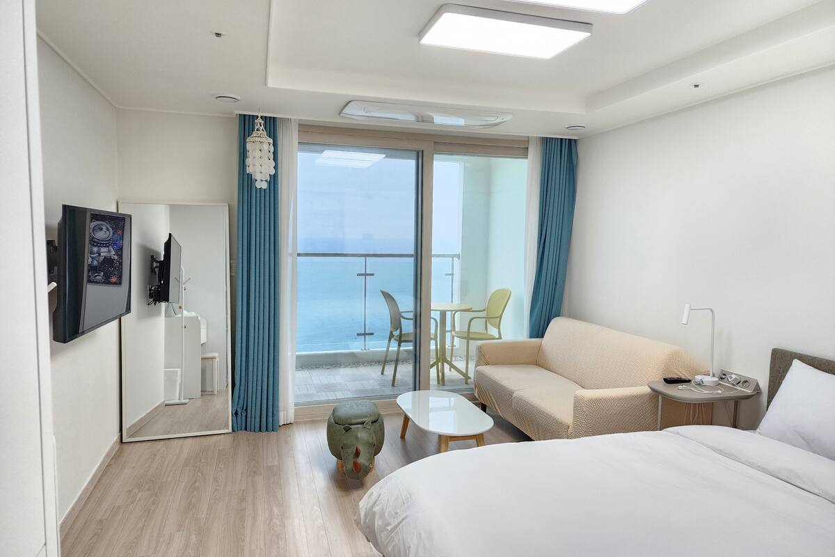 Quarto de hotel com uma cama, um sofá, uma mesinha e uma cadeira. Tem uma varanda com vista para o mar.