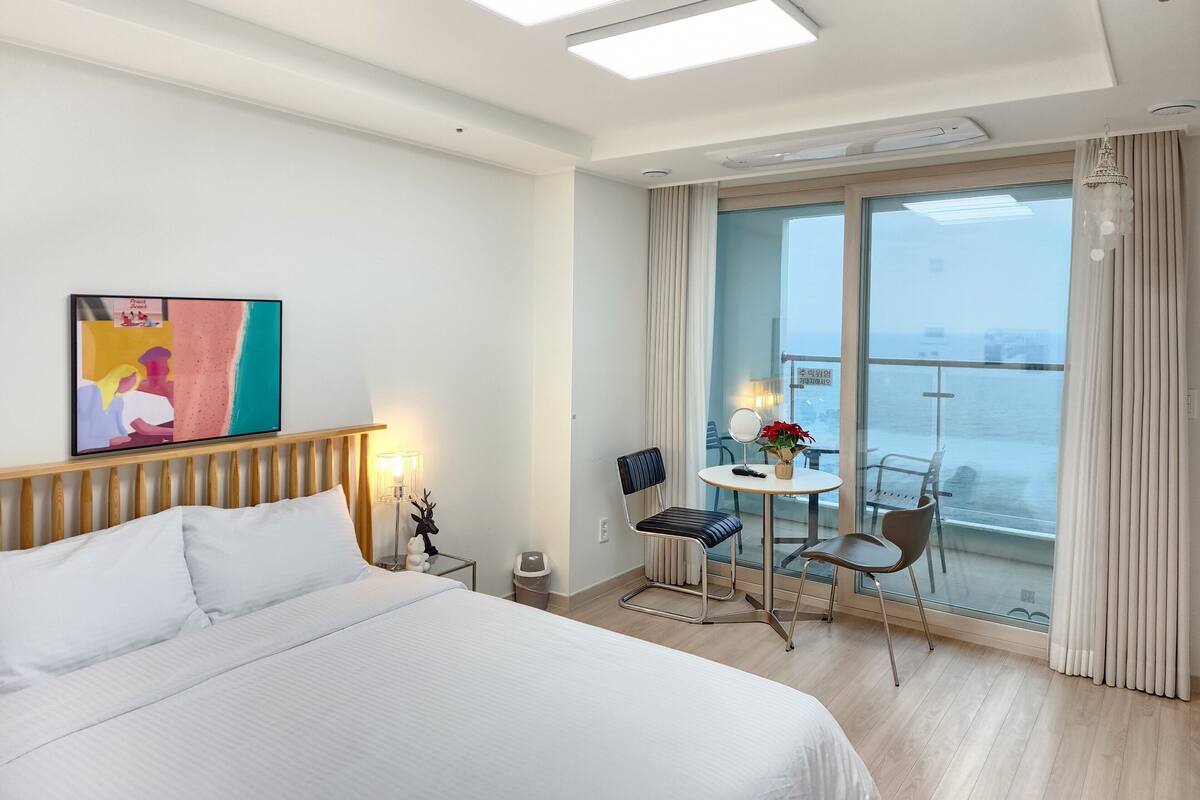 Quarto de hotel com uma cama, uma mesinha com uma cadeira, uma janela com vista para o mar e um quadro na parede.