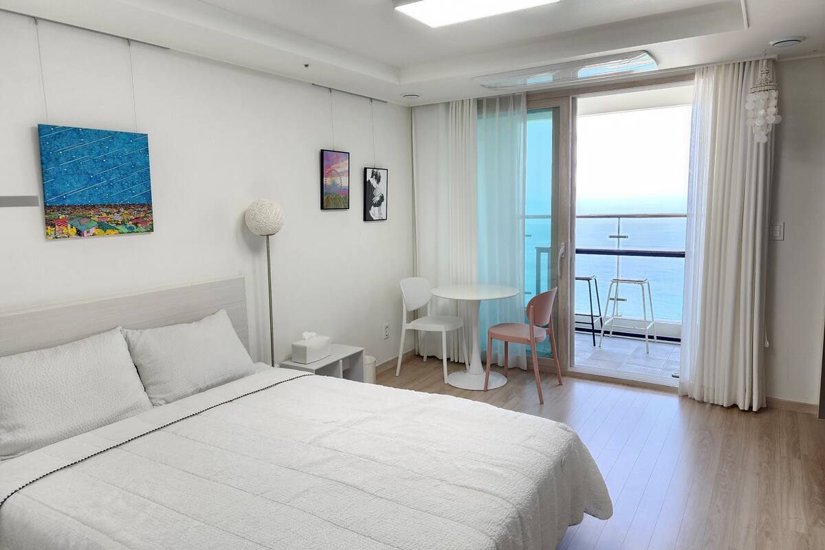 Quarto de hotel moderno com uma cama grande, uma mesa pequena com cadeiras, um abajur e uma varanda com vista para o mar.
