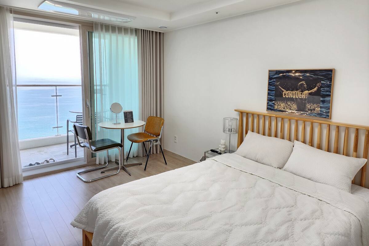 Quarto de hotel com cama, uma mesinha com cadeira, espelho, mesa de cabeceira e vista para o mar através de grandes janelas.