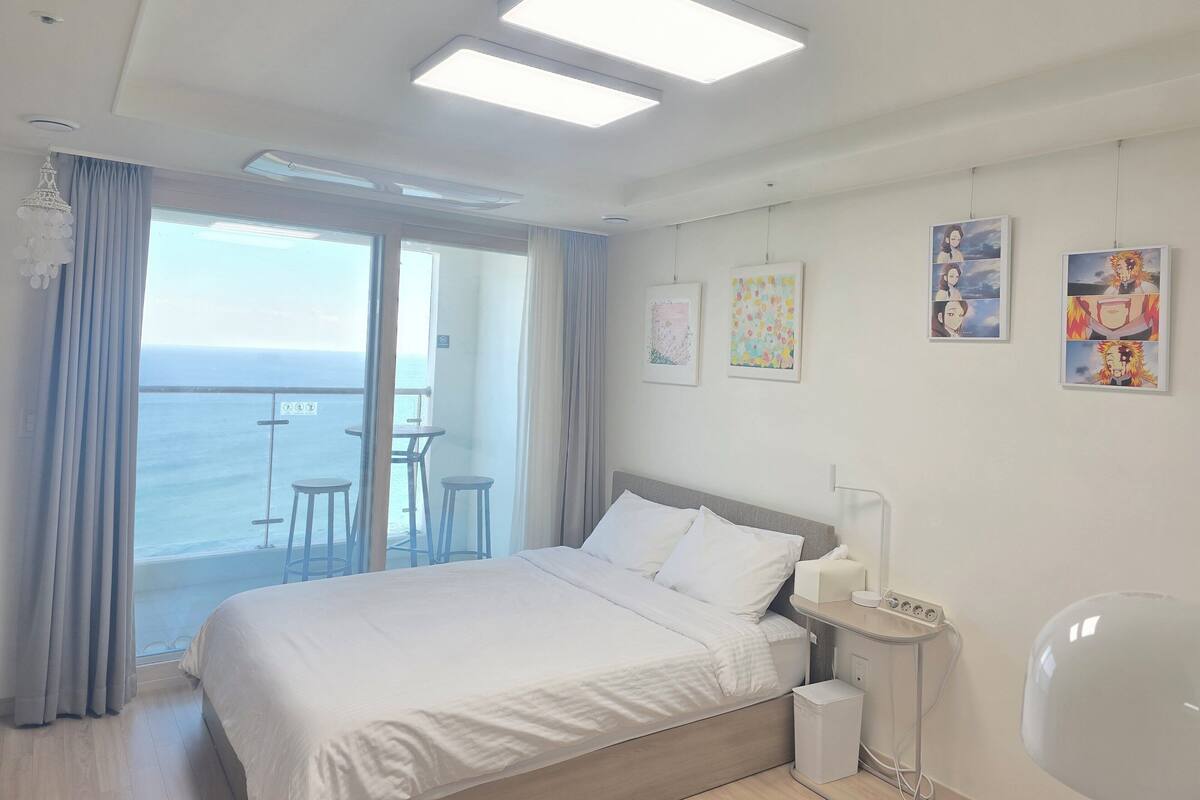 Quarto de hotel com cama, mesa de cabeceira, abajur e vista para o oceano através de uma porta de varanda.