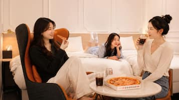 Quarto de hotel com cama, duas cadeiras, uma mesa com caixa de pizza e bebidas, e uma mesinha de cabeceira com abajur.