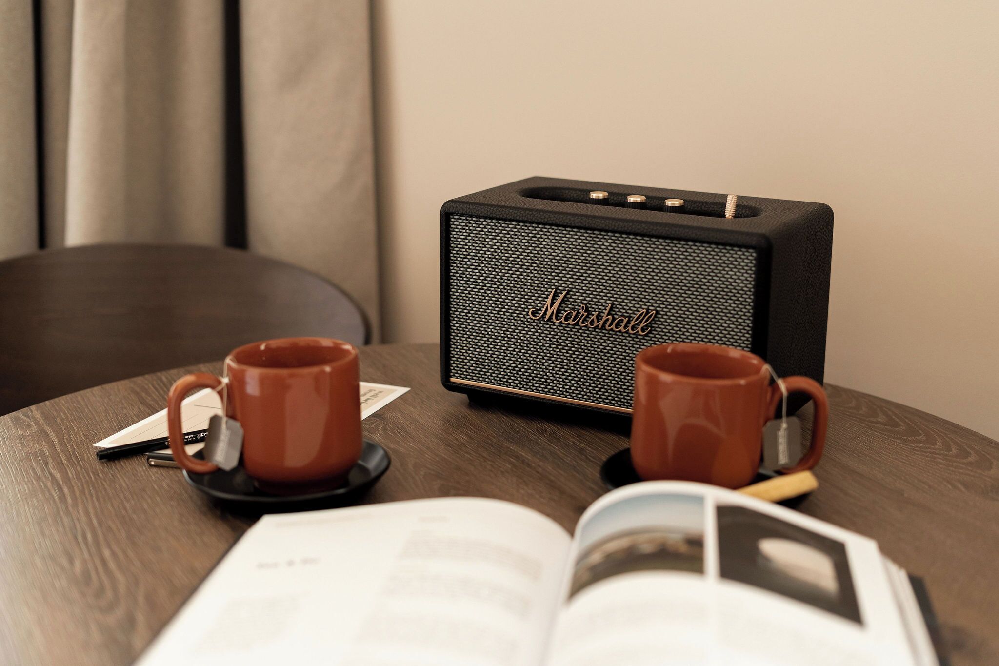 Uma mesa de madeira com um alto-falante Marshall, duas canecas, um livro e uma caneta.