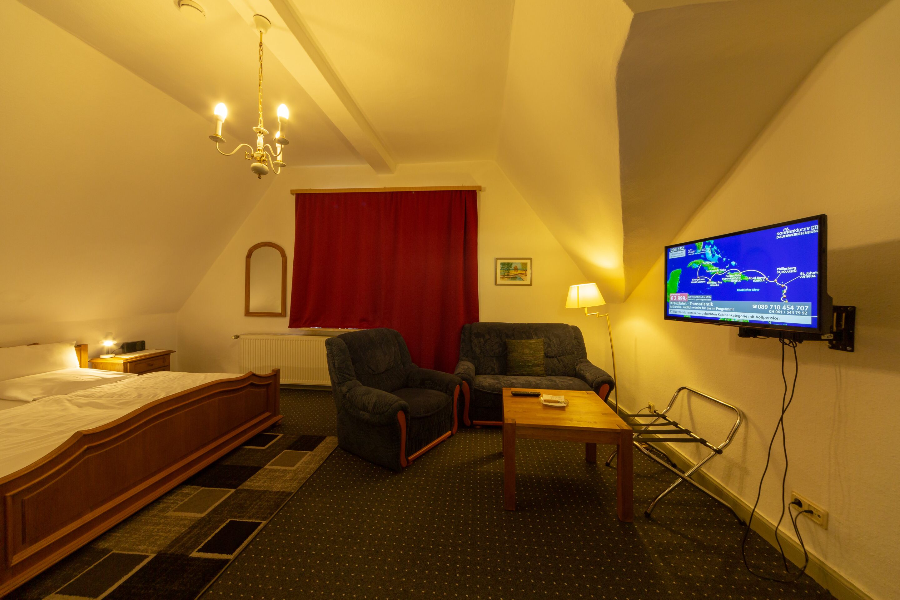 Quarto de hotel com cama, duas poltronas, uma mesa pequena, televisão e um lustre.