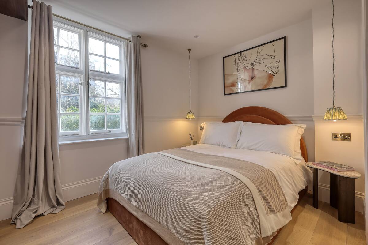 Quarto bem arrumado com cama grande, mesa de cabeceira com um livro, luminária pendente e janela com cortinas.