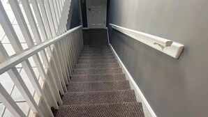 Uma escadaria com piso carpeteado, corrimãos brancos e uma porta no topo.