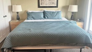 Uma cama bem arrumada com um cobreçado azul acolchoado, duas almofadas combinando, um desenho arquitetônico emoldurado na parede e duas luminárias de cabeceira de cada lado da cama.