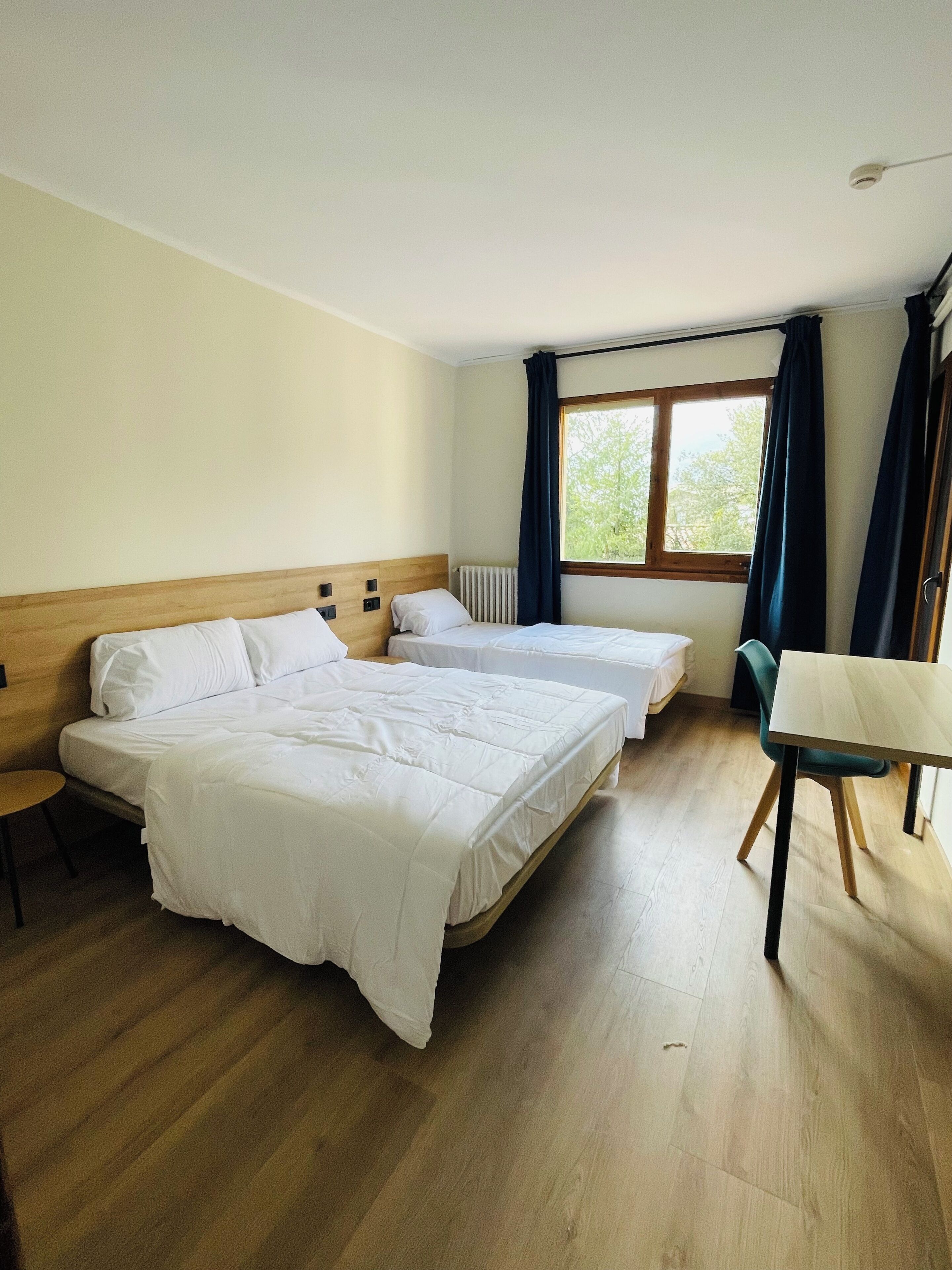 Quarto de hotel com duas camas, piso de madeira, janela com cortinas e uma mesa.