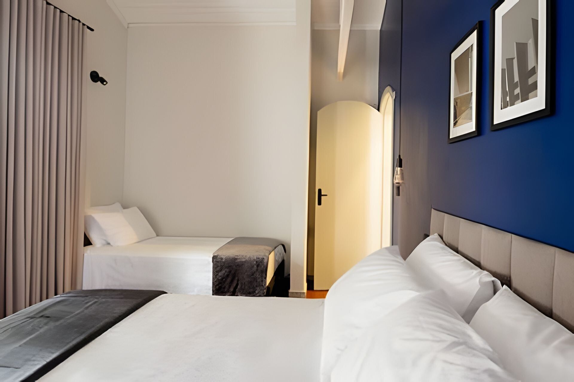 Quarto de hotel com duas camas, uma janela grande com cortinas, uma porta e uma parede com quadros.