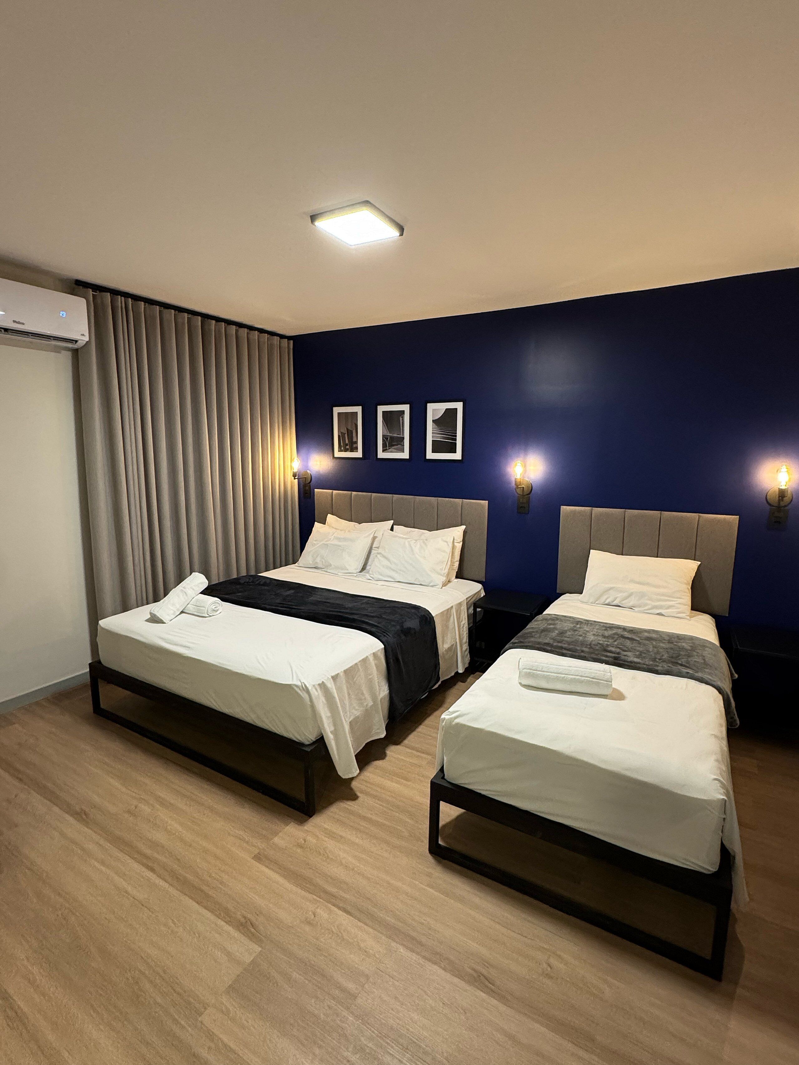 Quarto de hotel com duas camas, uma luminária de teto, ar condicionado e arandelas na parede.