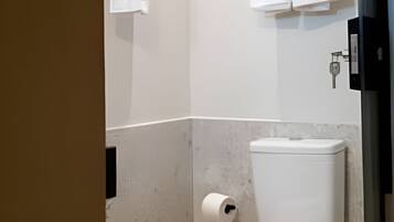 Uma casa de banho pequena e limpa, com sanita, um rolo de papel higiénico e duas toalhas brancas penduradas na parede.