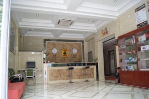 Uma área de recepção de hotel com piso de mármore, balcão de atendimento, sala de espera com cadeiras e um expositor com objetos.