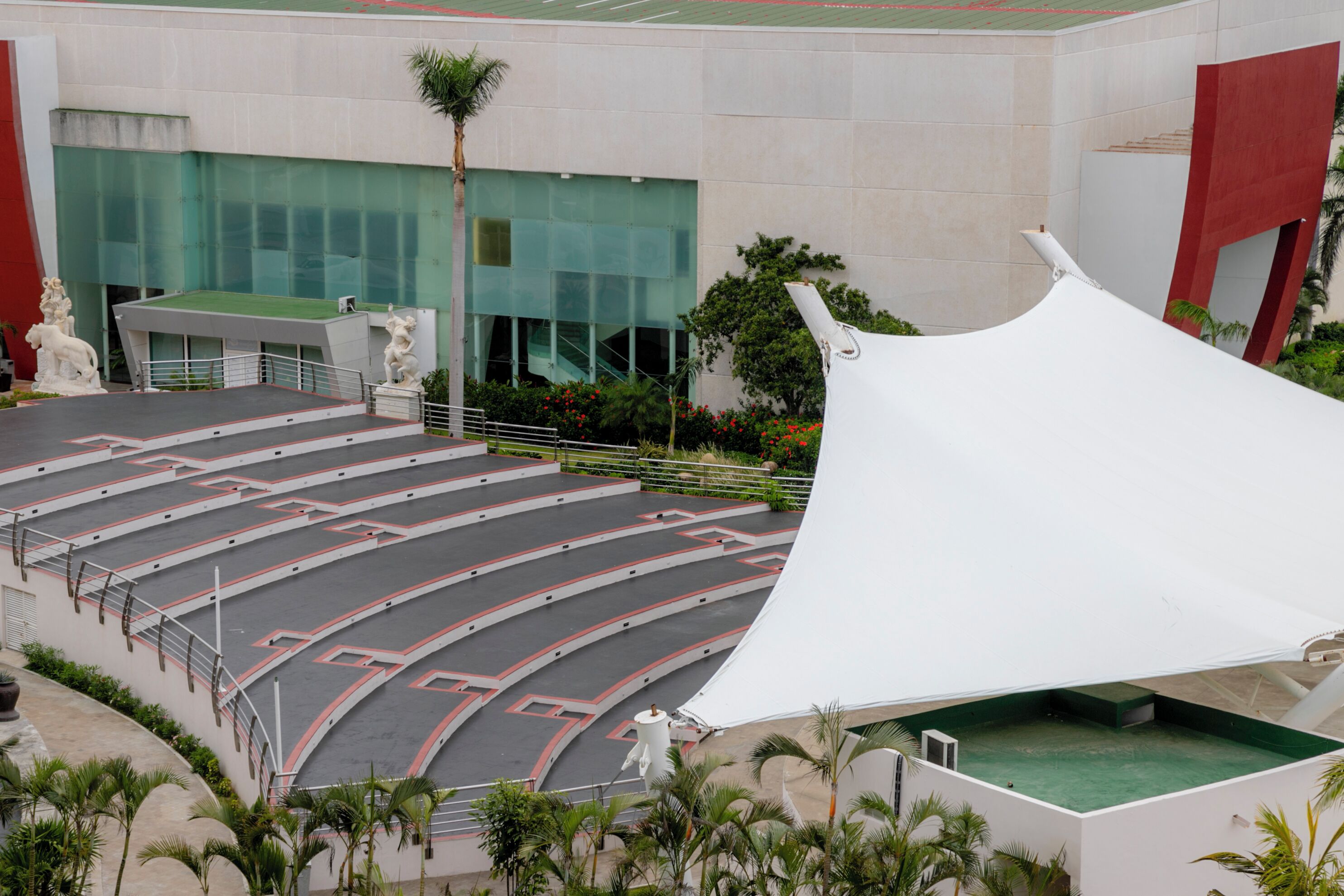 Um anfiteatro ao ar livre com assentos em níveis, uma cobertura branca e um edifício com janelas de vidro e detalhes vermelhos ao fundo.
